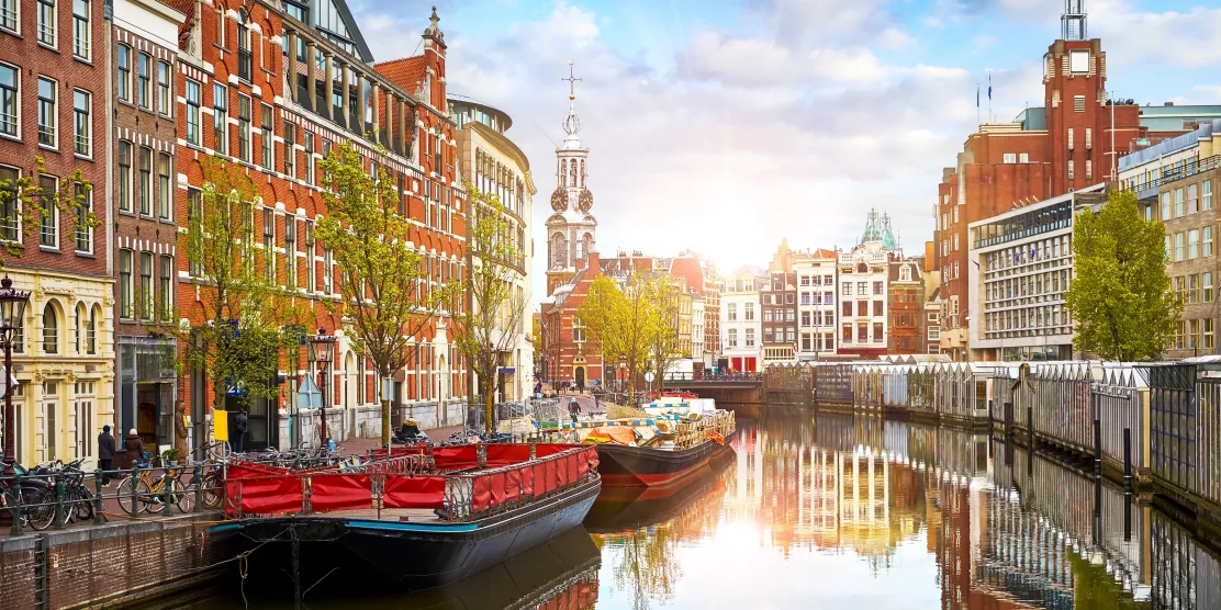 View of an Amsterdam canal in the Netherlands, showing traditional houseboats moored beside historic gabled buildings and the Munttoren tower, with bicycles parked along the waterside.