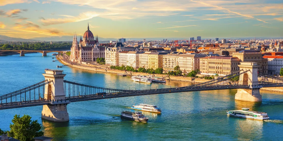 Aerial view of Budapest, Hungary at sunset, showing the Chain Bridge crossing the Danube River with sightseeing boats, and the Hungarian Parliament Building's domed rooftop in the background.