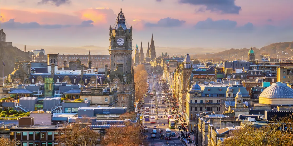 Aerial view of Edinburgh, Scotland at sunset, showing the Balmoral Clock Tower and Princes Street bustling with buses, cars and pedestrians, with Edinburgh Castle visible on the hill in the background.