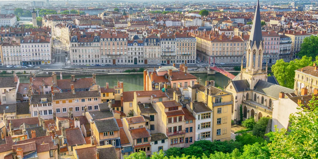 Aerial view of Lyon, France, showing dense rows of historic buildings with terracotta roofs along the Saône River, with a church spire in the foreground and the city skyline stretching into the distance.
