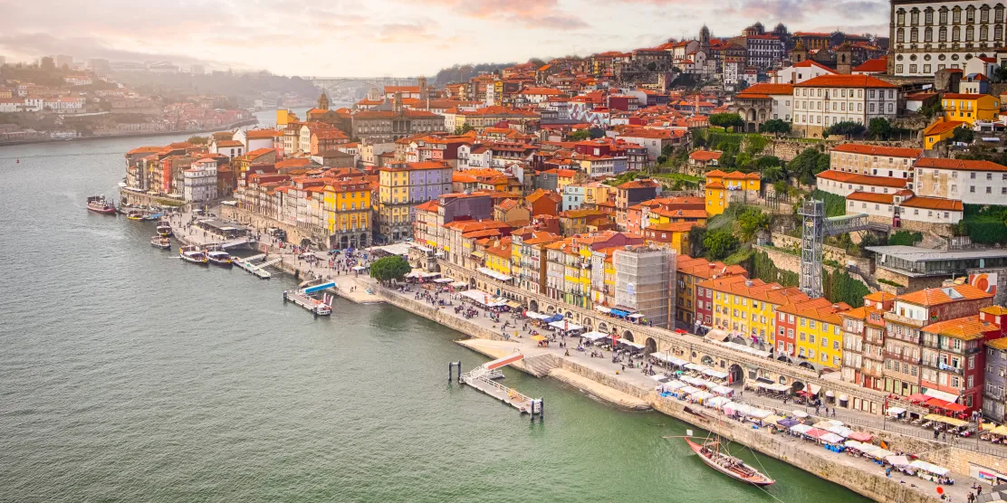 Aerial view of Porto, Portugal at sunset, showing colourful riverside buildings with orange terracotta roofs along the Douro River, with traditional rabelo boats moored at the waterfront promenade