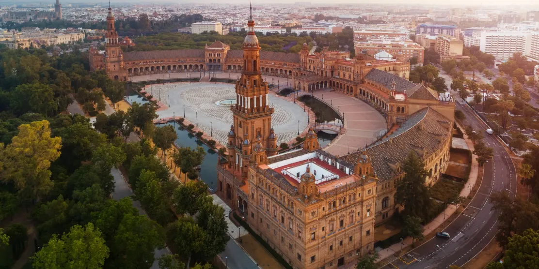 Aerial view of the Plaza de España in Seville, Spain at sunrise, showing its curved brick facade, ornate tower and central plaza with a fountain, surrounded by trees.