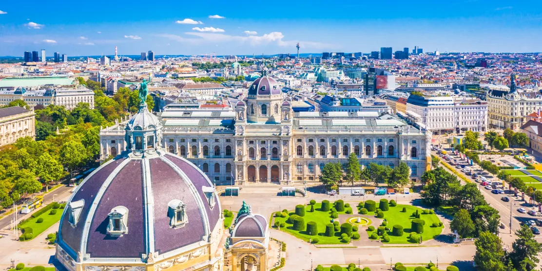 Aerial view of the Natural History Museum in Vienna, Austria, showing its ornate dome and facade overlooking manicured gardens, with the modern city skyline in the distance.