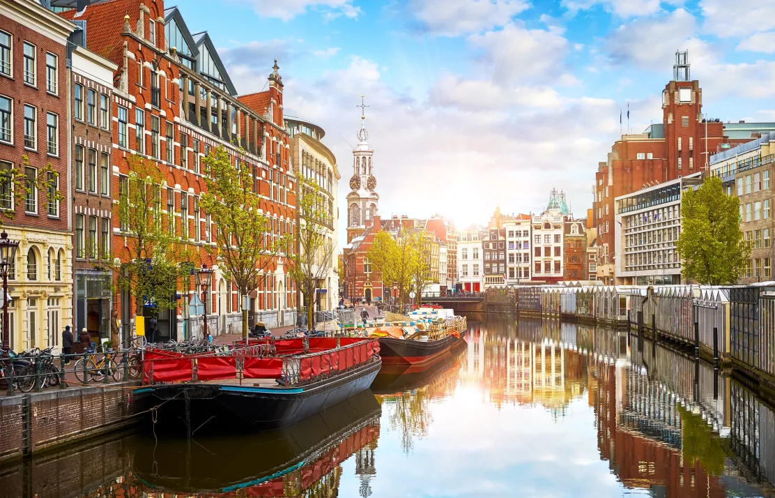 View of an Amsterdam canal in the Netherlands, showing traditional houseboats moored beside historic gabled buildings and the Munttoren tower, with bicycles parked along the waterside.