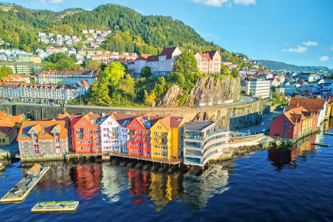 Aerial view of Bergen, Norway, showing colourful wooden waterfront buildings in red, yellow and white built on stilts over the harbour, backed by a forested hillside with clifftop houses.