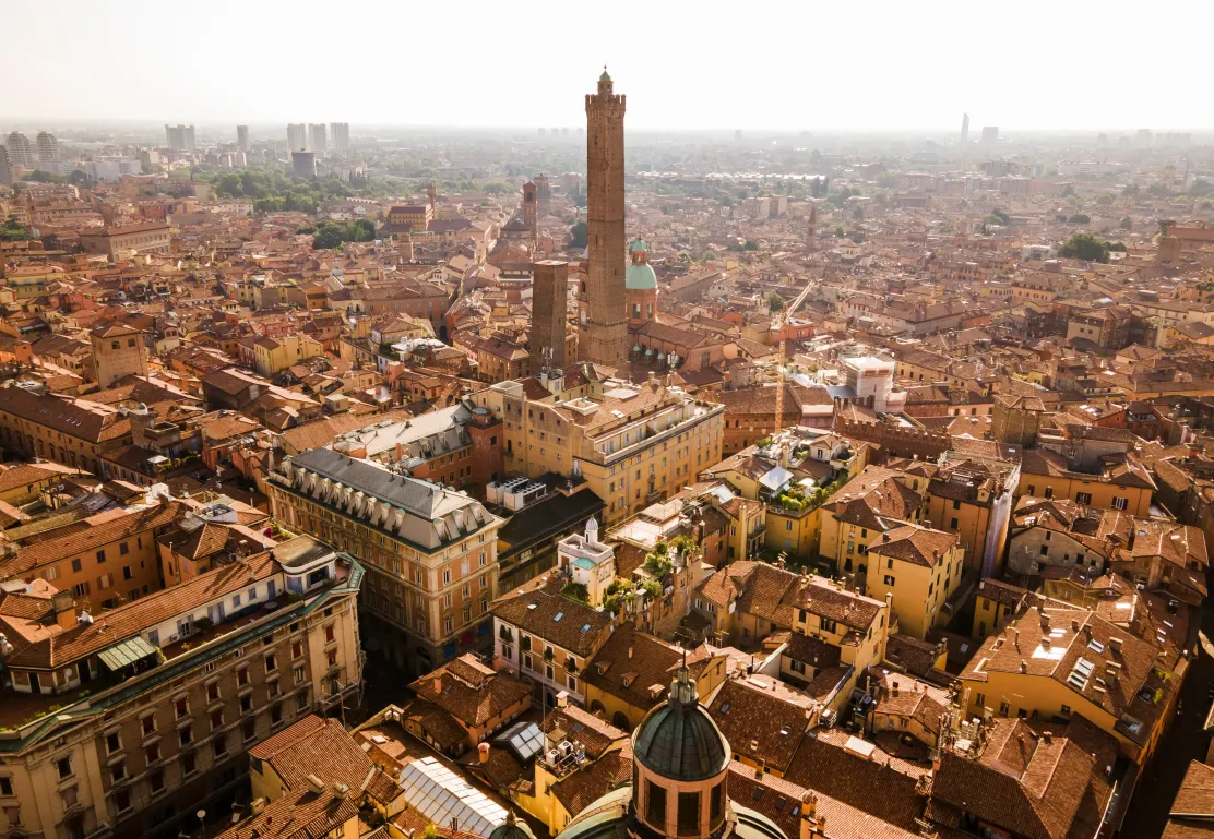 Aerial view of Bologna, Italy, showing the city's iconic leaning medieval towers rising above a dense sea of orange terracotta rooftops.