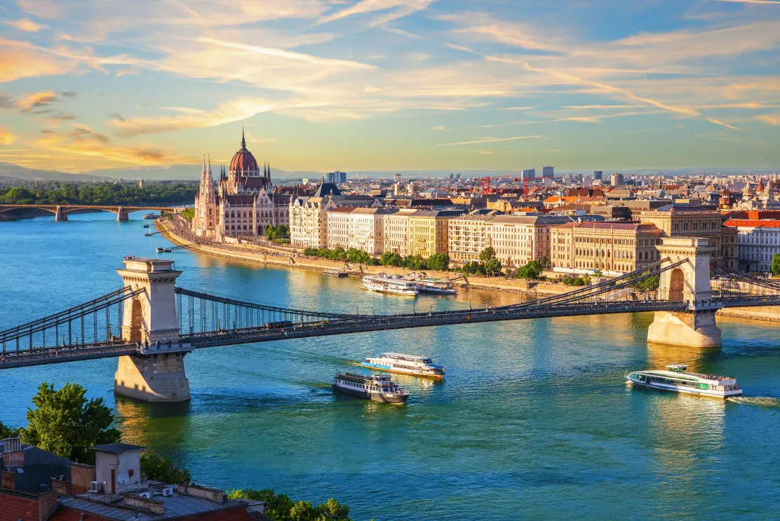 Aerial view of Budapest, Hungary at sunset, showing the Chain Bridge crossing the Danube River with sightseeing boats, and the Hungarian Parliament Building's domed rooftop in the background.
