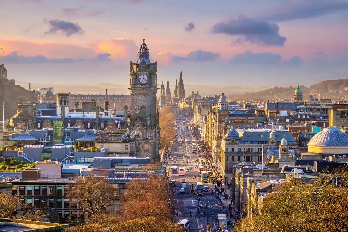 Aerial view of Edinburgh, Scotland at sunset, showing the Balmoral Clock Tower and Princes Street bustling with buses, cars and pedestrians, with Edinburgh Castle visible on the hill in the background.