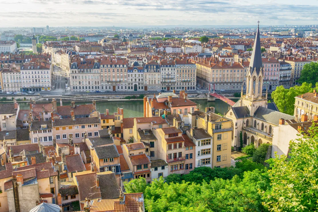Aerial view of Lyon, France, showing dense rows of historic buildings with terracotta roofs along the Saône River, with a church spire in the foreground and the city skyline stretching into the distance.