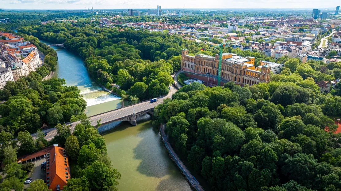 Aerial view of Munich, Germany, showing the Maximilianeum building surrounded by lush green forest along the Isar River, with a bridge crossing the water in the foreground.
