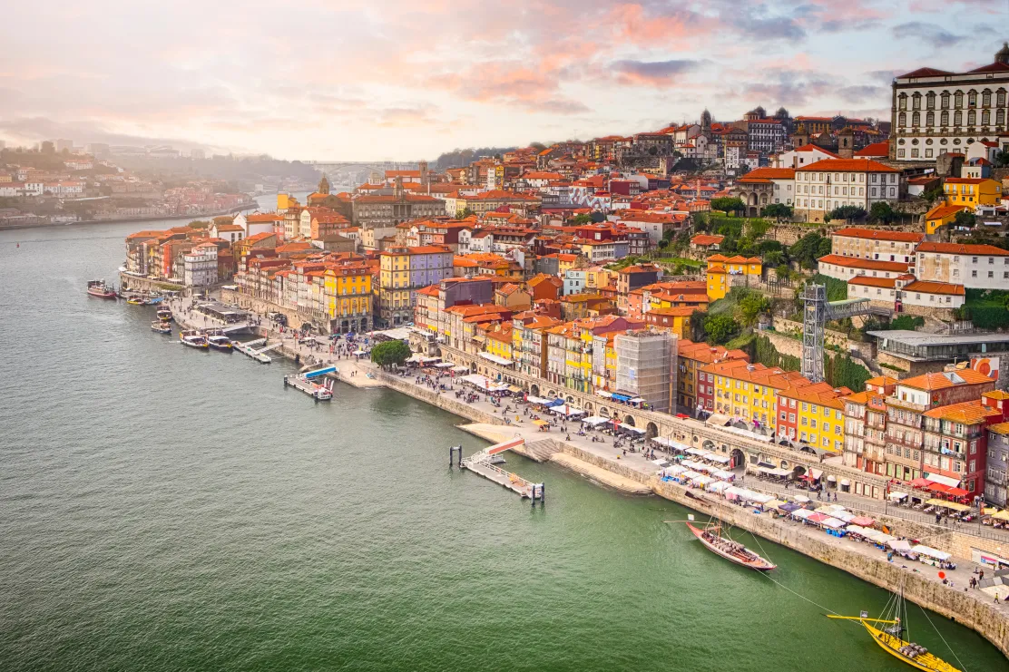 Aerial view of Porto, Portugal at sunset, showing colourful riverside buildings with orange terracotta roofs along the Douro River, with traditional rabelo boats moored at the waterfront promenade