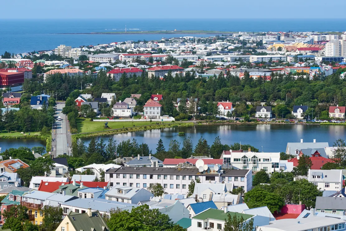 Aerial view of Reykjavik, Iceland, showing a colourful residential neighbourhood with red and blue rooftops surrounding a city pond, with the ocean visible in the background.