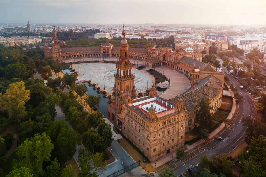 Aerial view of the Plaza de España in Seville, Spain at sunrise, showing its curved brick facade, ornate tower and central plaza with a fountain, surrounded by trees.