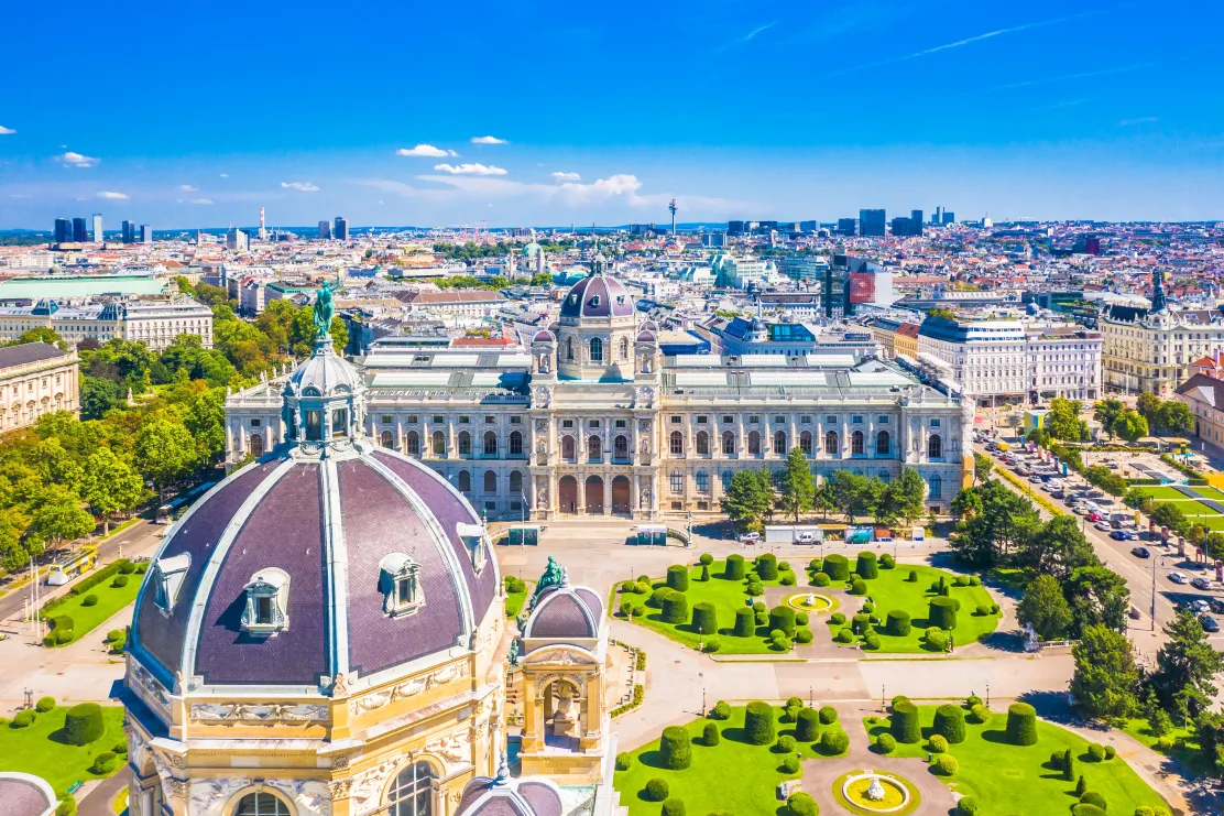 Aerial view of the Natural History Museum in Vienna, Austria, showing its ornate dome and facade overlooking manicured gardens, with the modern city skyline in the distance.
