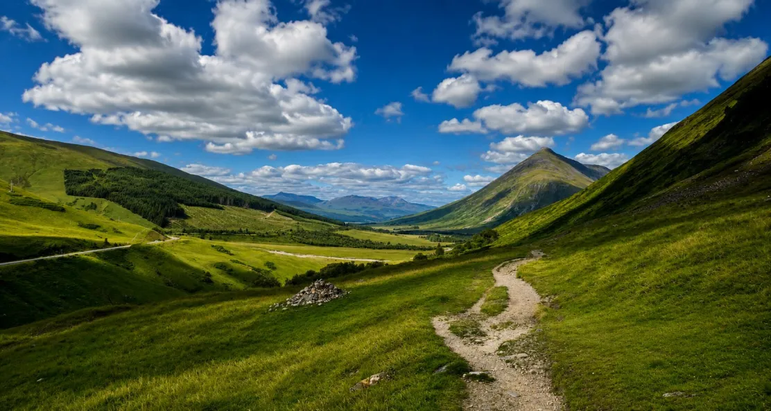 West Highland Way hiking trail winding through a vast Scottish glen with green rolling hills and historic stone ruins under a moody sky.