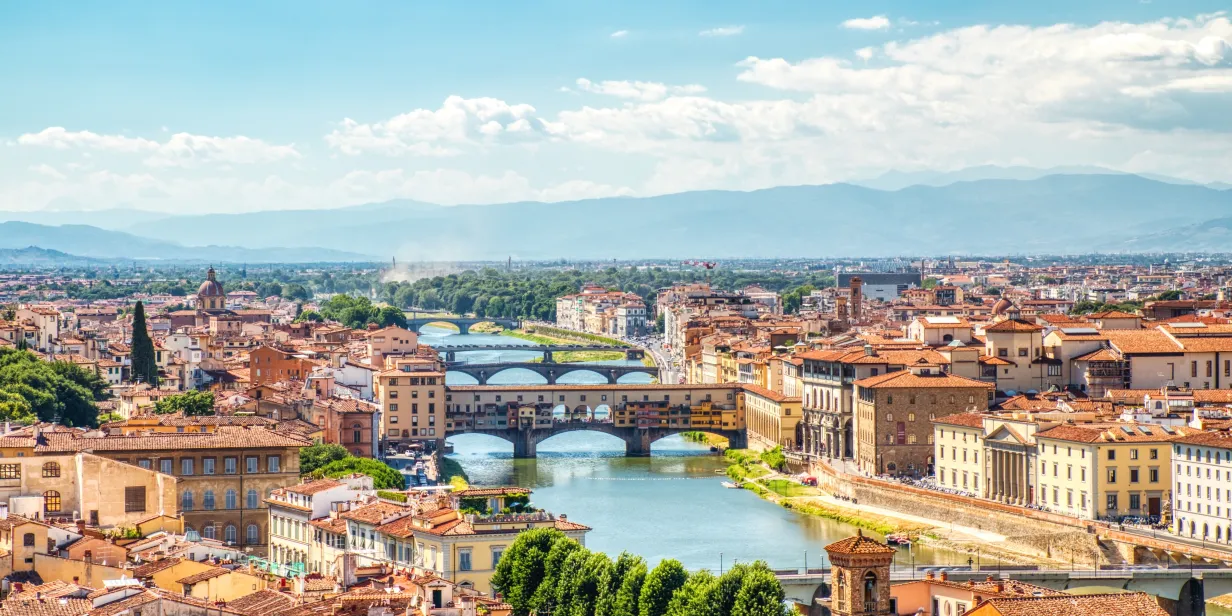 Aerial view of Florence, Italy, showing the Ponte Vecchio bridge spanning the Arno River, surrounded by terracotta rooftops with the Tuscan hills in the background.